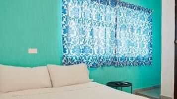 Quarto com cama, janela com cortinas estampadas, ar condicionado e uma mesinha de cabeceira.