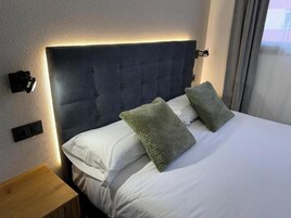 Cama bem arrumada com cabeceira estofada, dois travesseiros e mesa de cabeceira com abajur.