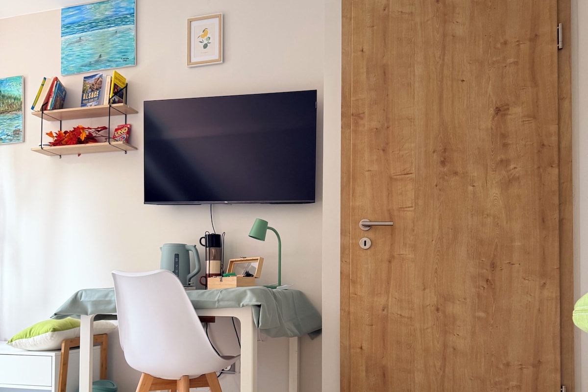 Quarto com cama, escrivaninha, cadeira, televisão e porta de madeira.
