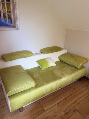 Cama com almofadas verdes e cabeceira branca num quarto com soalho de madeira.