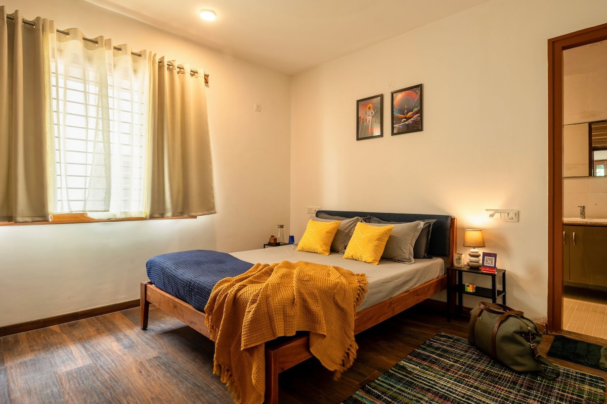 Quarto com cama, mesa de cabeceira, luminária, janela com cortinas e um quadro na parede.