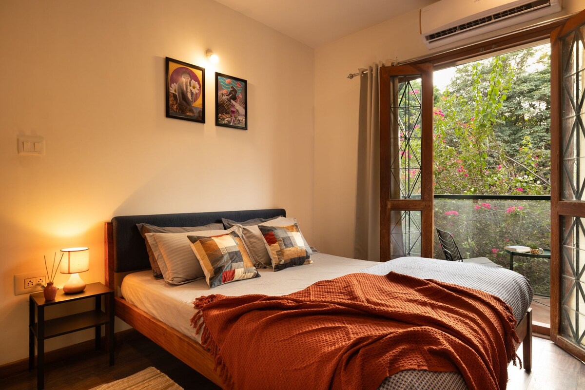 Quarto com cama, almofadas, abajur e janela com vista para áreas verdes.