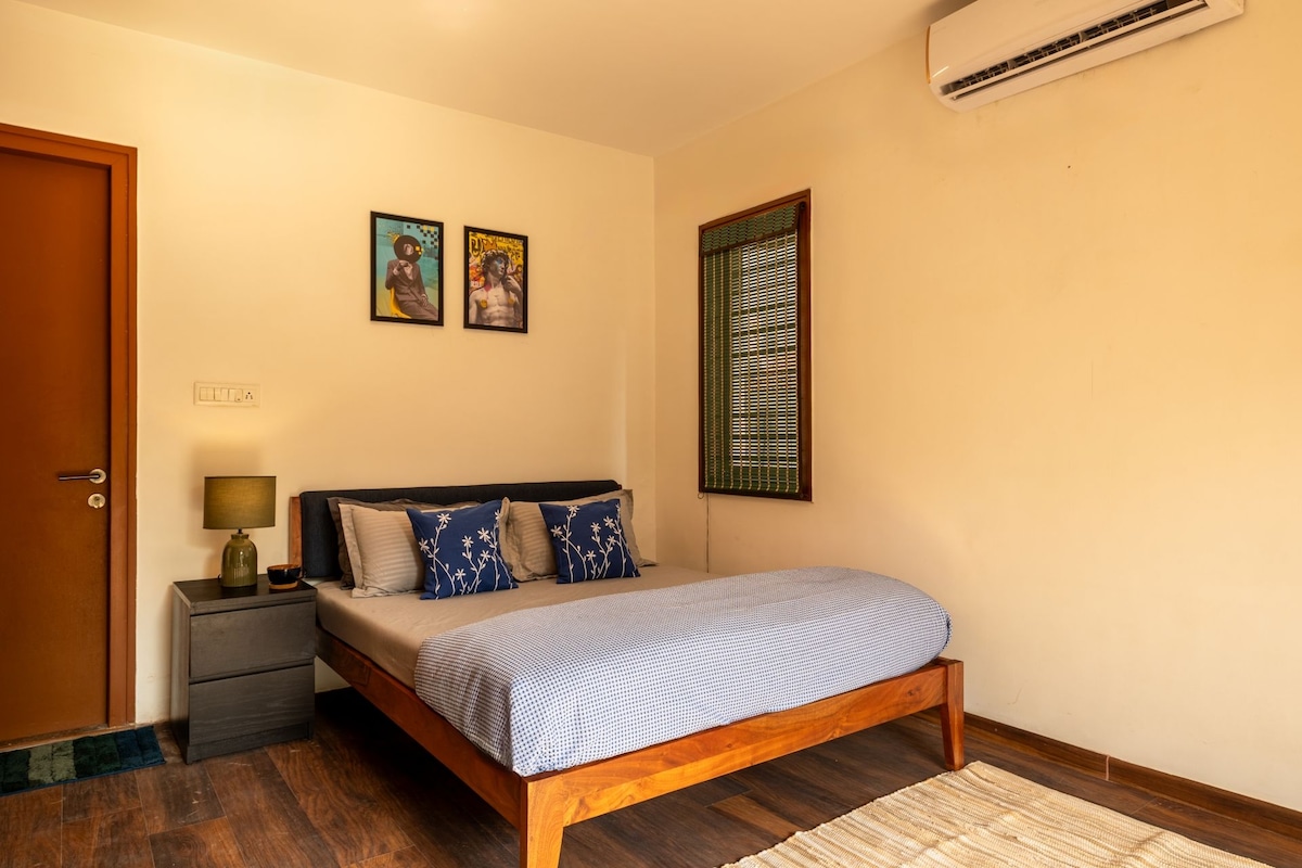 Quarto com cama de madeira, mesa de cabeceira, luminária, ar-condicionado fixado na parede e duas fotos emolduradas.