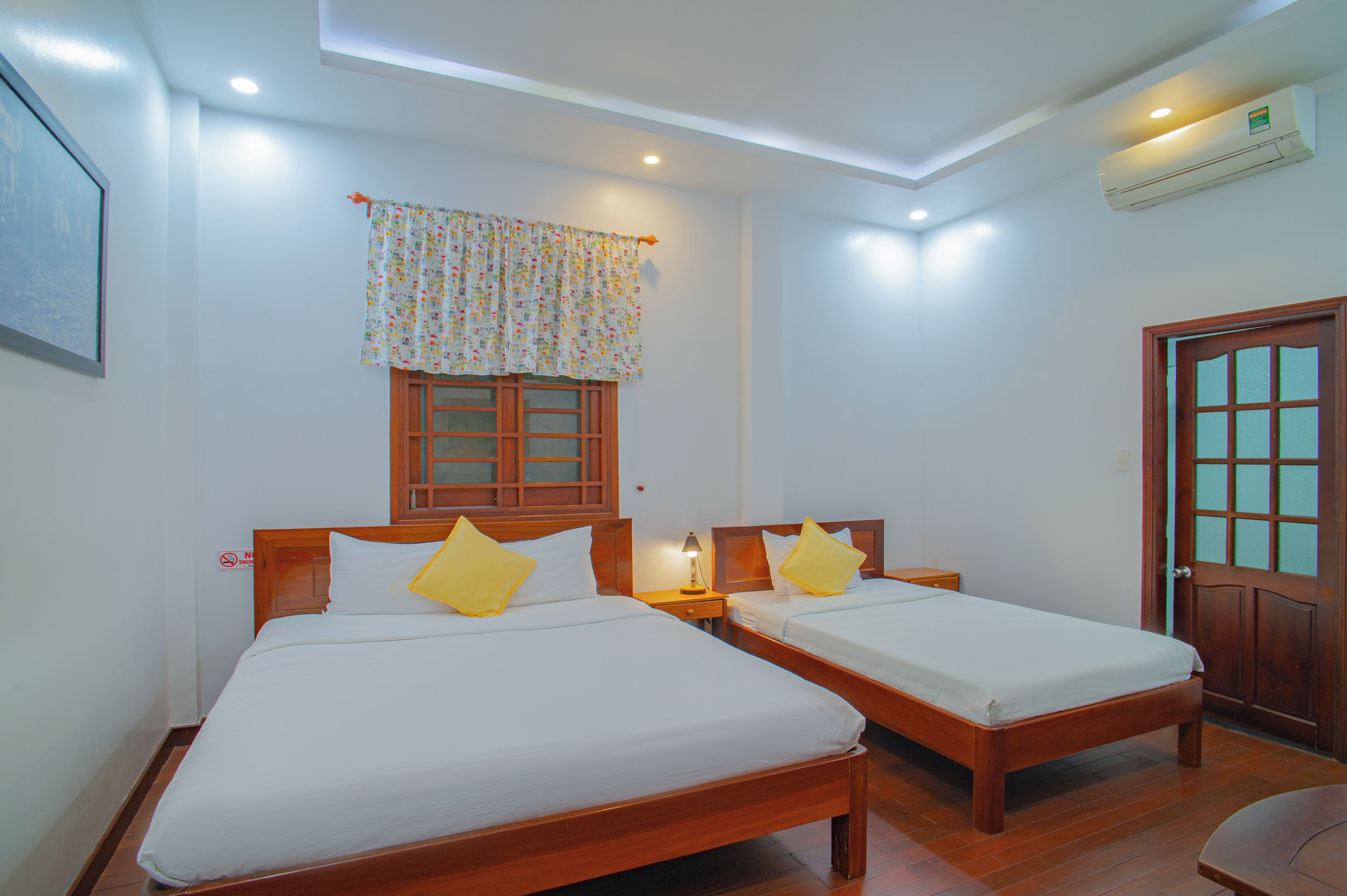 Quarto de hotel com duas camas de madeira, janela com cortina estampada, porta com painéis de vidro e ar condicionado na parede.