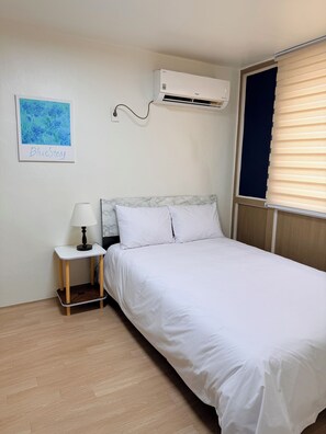 Quarto com cama, mesa de cabeceira com abajur, ar-condicionado e janela com persianas.