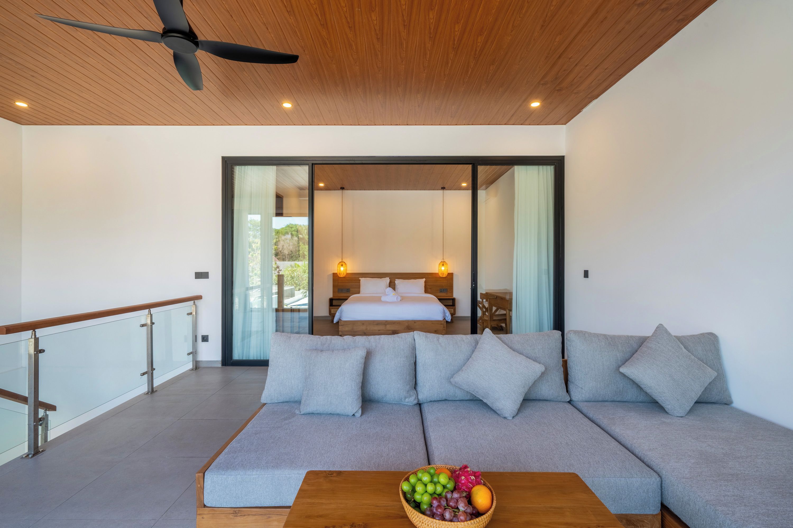 Quarto de hotel moderno com um sofá grande, mesa de centro de madeira com frutas e uma varanda com guarda-corpo de vidro.
