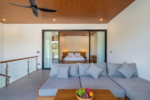 Quarto de hotel moderno com um sofá grande, mesa de centro de madeira com frutas e uma varanda com guarda-corpo de vidro.