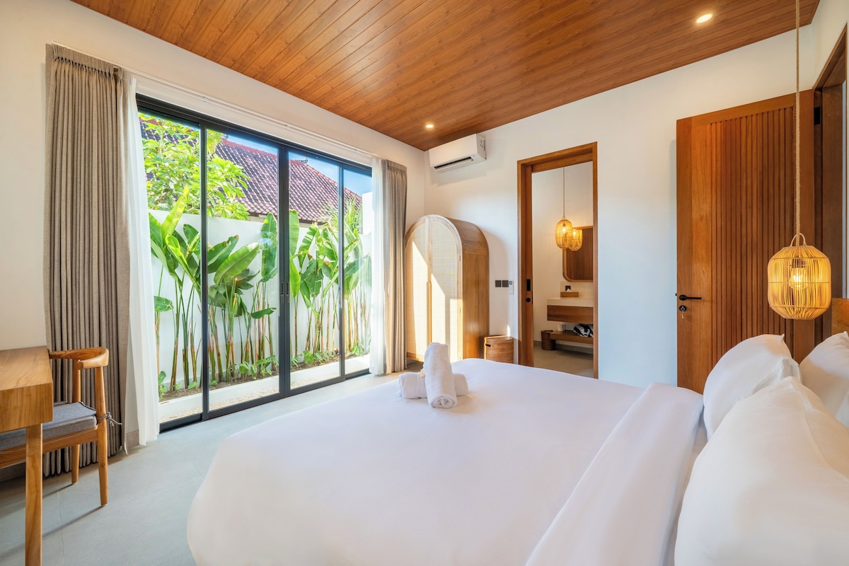 Quarto de hotel com uma cama grande, teto de madeira, porta de vidro deslizante para um jardim e uma pequena mesa com uma cadeira.