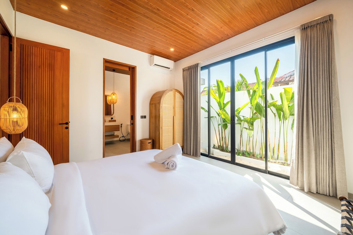 Quarto de hotel com cama grande, teto de madeira e vista para o verde pela janela.