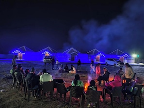 Um grupo de pessoas sentadas ao redor de uma fogueira em área aberta, com vários edifícios ao fundo.