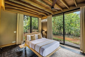 Quarto com uma cama grande, ventilador de teto, aparelho de ar condicionado e vista para um jardim exuberante através de grandes janelas.