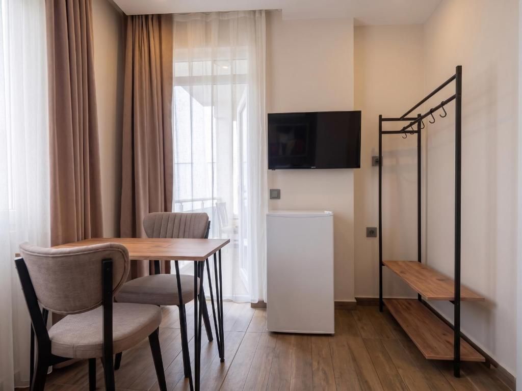 Quarto compacto com mesa de jantar, cadeiras, televisão fixada na parede, geladeira branca e estrutura de cama de metal com prateleiras.