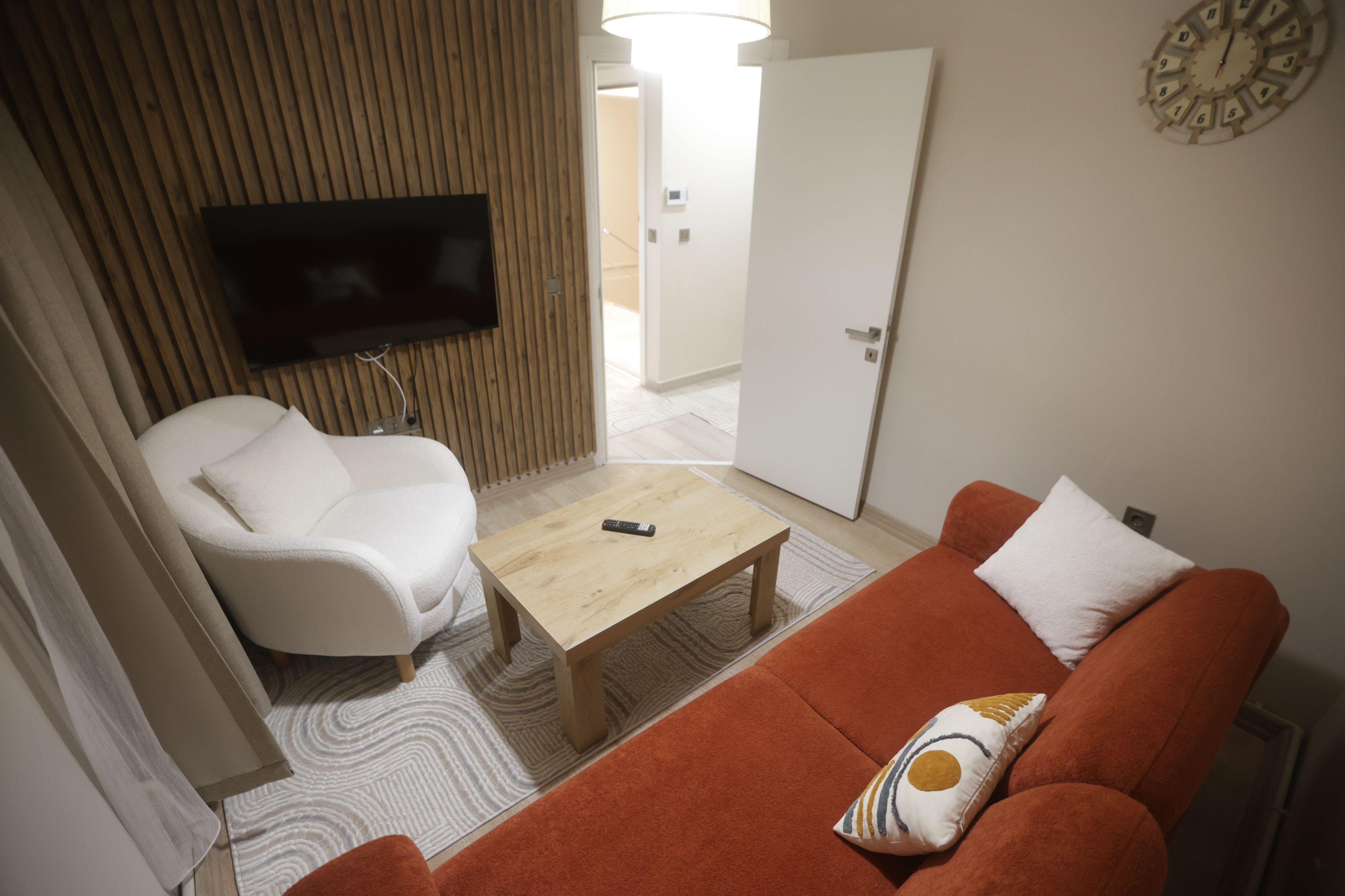Uma sala de estar com um sofá branco, um sofá laranja, uma mesa de centro de madeira e uma televisão fixada em uma parede de madeira.