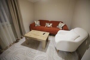 Uma sala de estar com sofá, poltrona branca, mesa de centro e um controle remoto sobre a mesa.