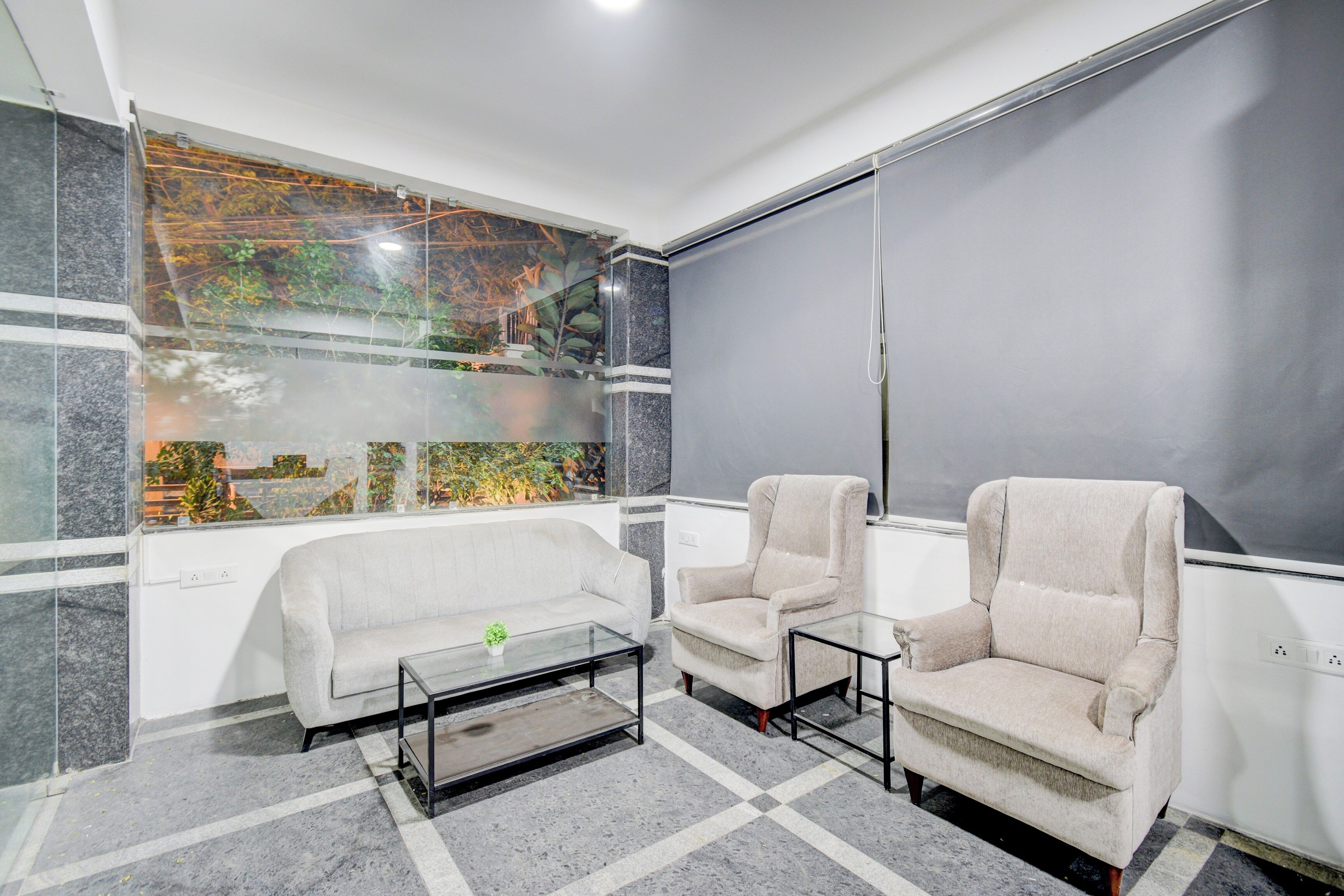 Quarto moderno com sofá cinzento, duas poltronas, mesa de centro de vidro e uma janela grande com vista para o verde.