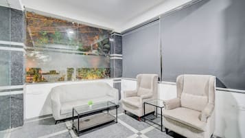 Quarto moderno com sofá cinzento, duas poltronas, mesa de centro de vidro e uma janela grande com vista para o verde.