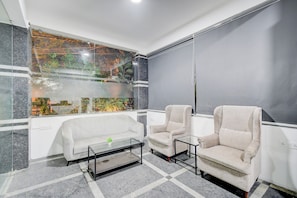Quarto moderno com sofá cinzento, duas poltronas, mesa de centro de vidro e uma janela grande com vista para o verde.