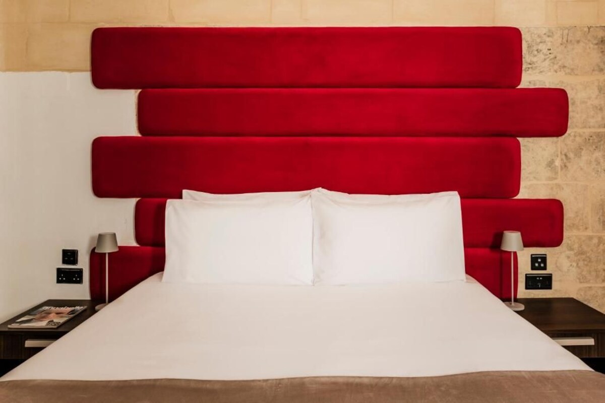 Quarto de hotel com cabeceira estofada vermelha, roupa de cama branca e duas mesas de cabeceira com luminárias.