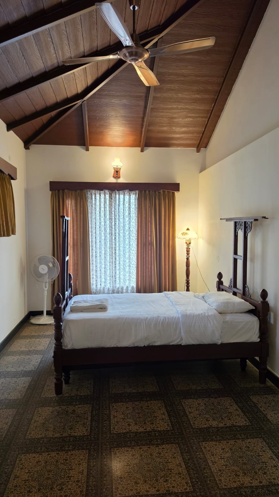 Quarto com teto de madeira, cama, ventilador e janela com cortinas.