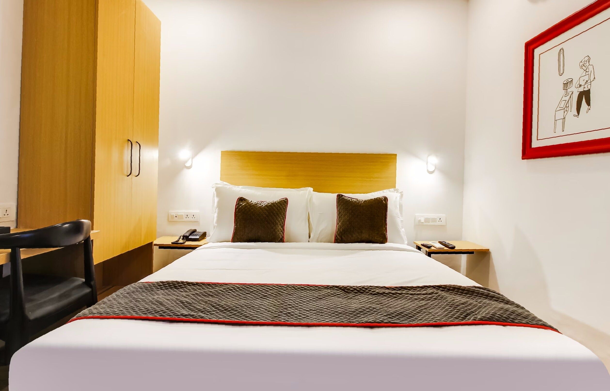 Quarto de hotel com cama, mesas de cabeceira, escrivaninha, cadeira e uma obra de arte na parede.