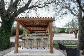 Uma pérgola de madeira com telhado de telhas, paredes de pedra e assentos acolchoados verdes.