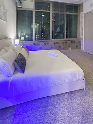 Uma cama bem arrumada com cabeceira, uma janela com persianas e piso coberto por carpete.