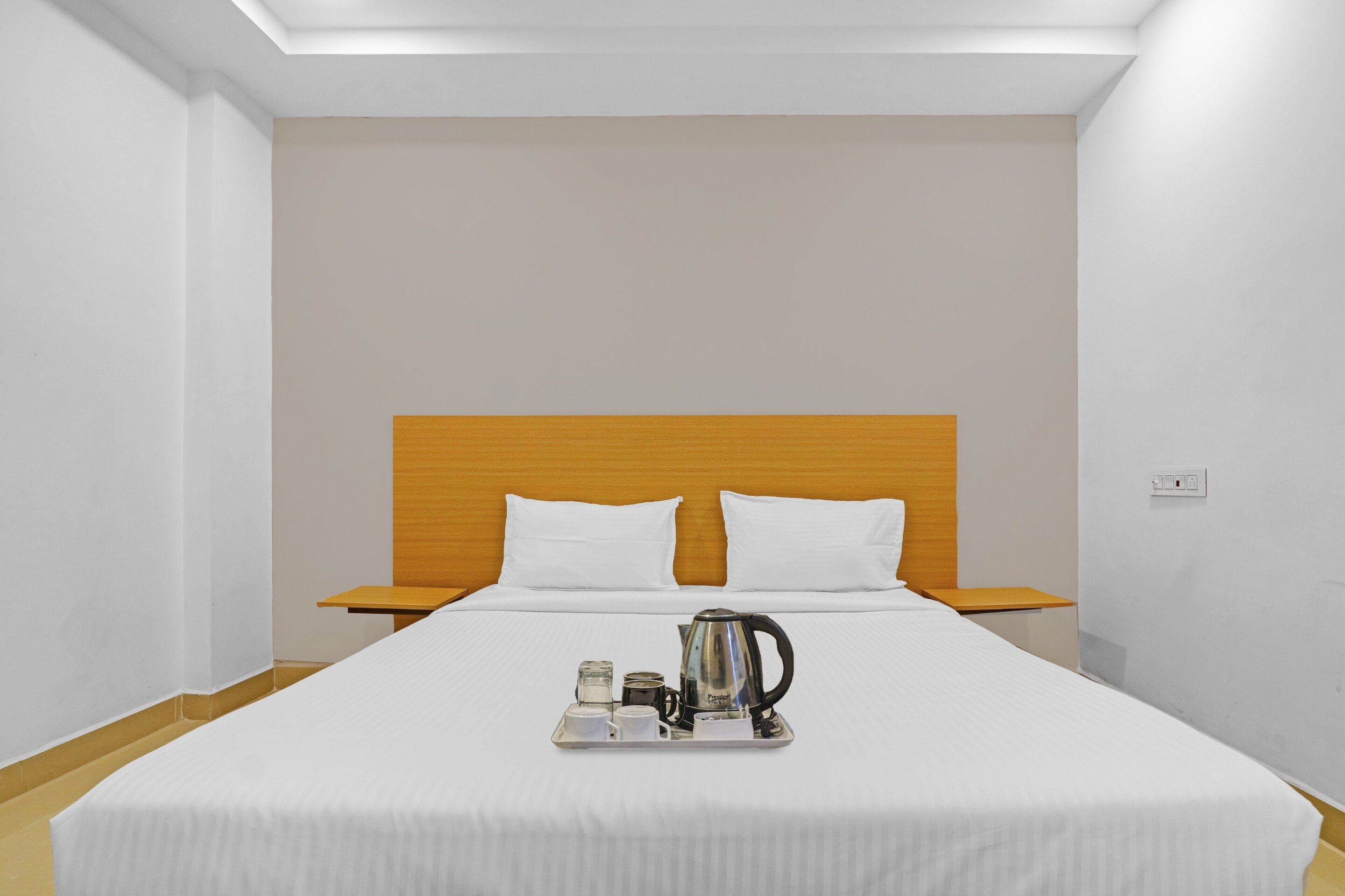 Quarto de hotel com uma cama de solteiro, cabeceira de madeira, duas almofadas brancas, um suporte com chaleira e xícaras, e uma mesa de cabeceira com abajur.
