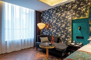 Quarto de hotel com sofá, mesinha redonda, luminária decorativa e cama com colcha estampada.