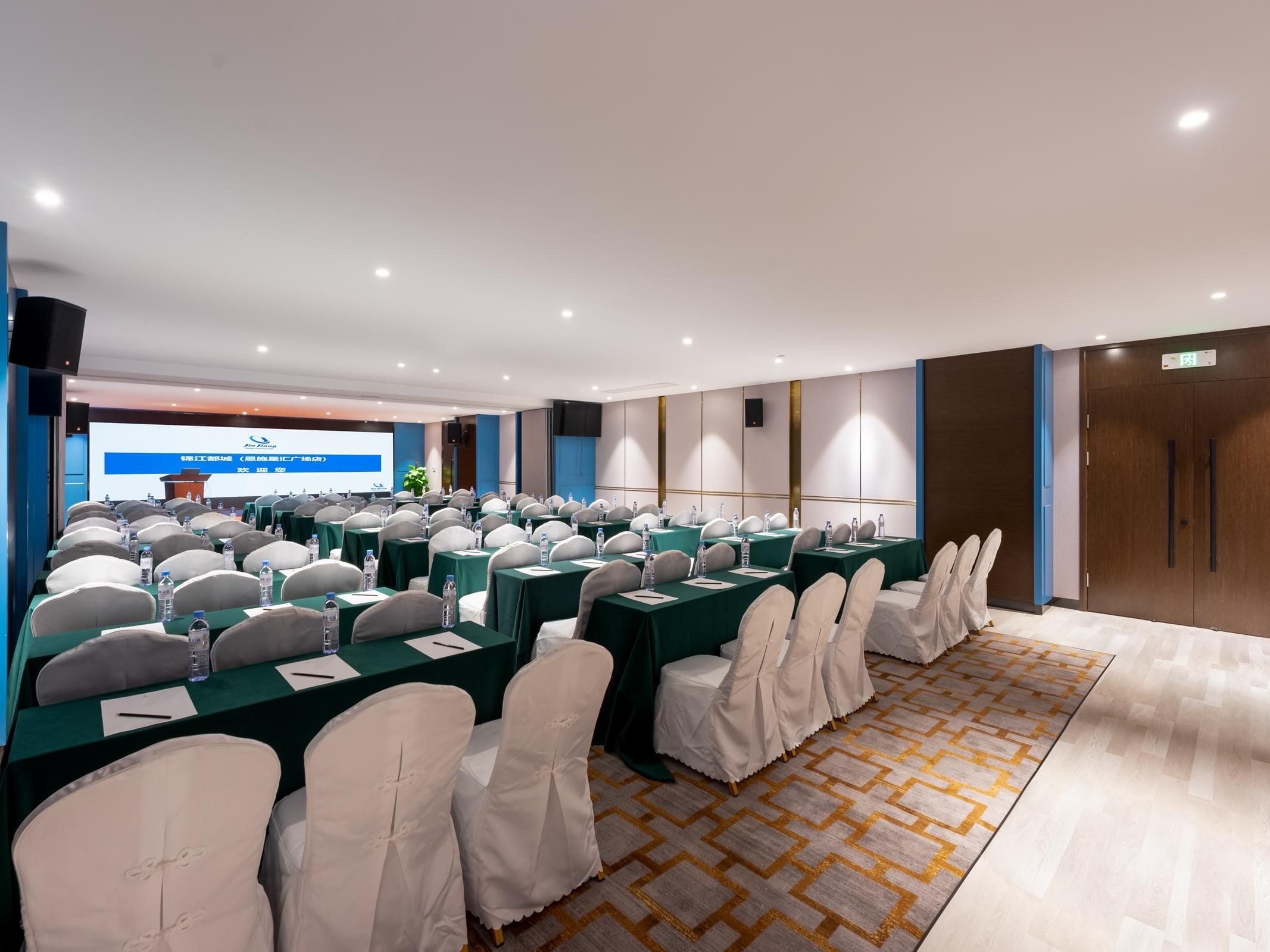 Sala de conferências com fileiras de mesas, cadeiras brancas e uma tela de apresentação.