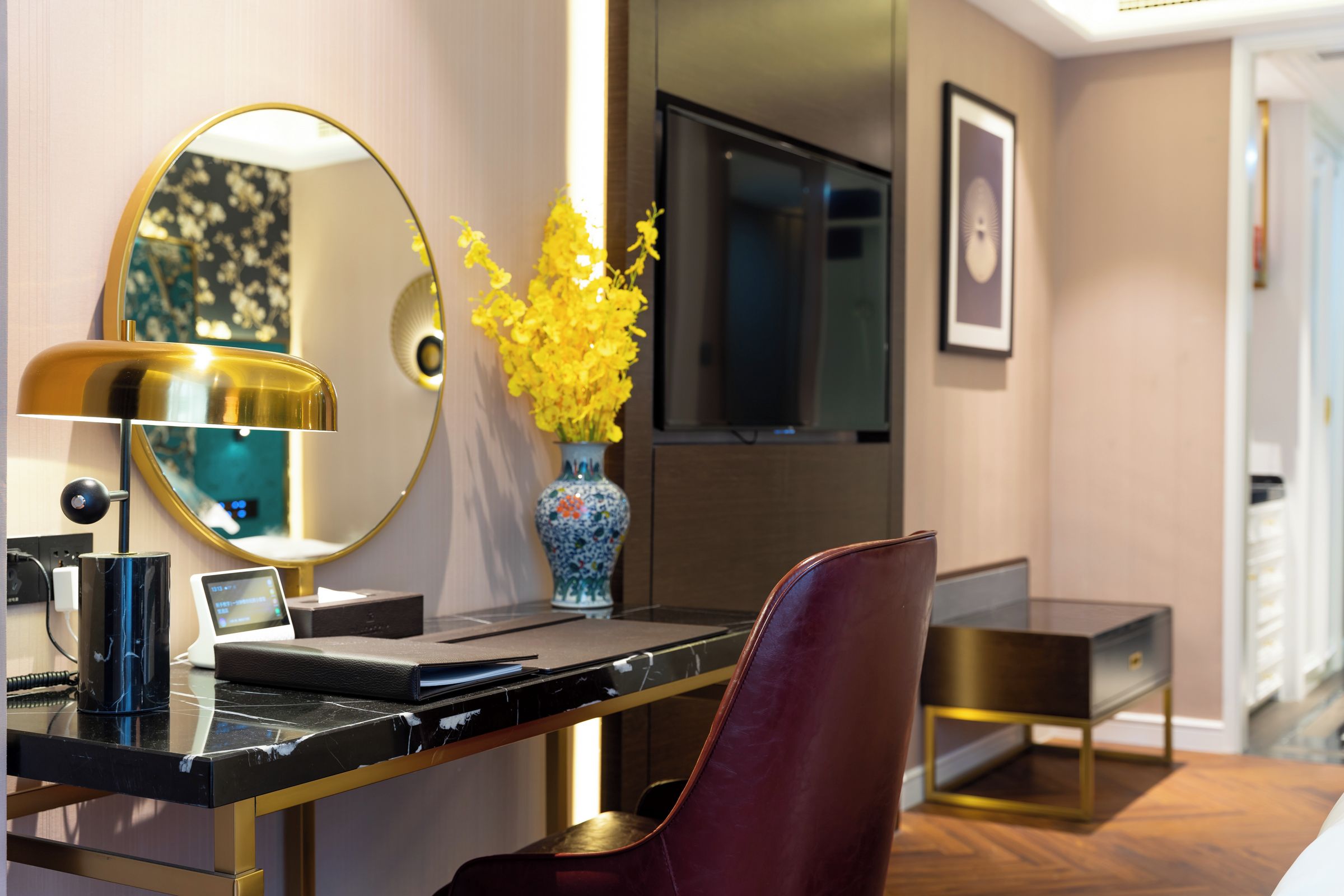 Quarto de hotel moderno com uma mesa de escritório, um espelho redondo grande, um vaso com flores amarelas, uma televisão e um quadro na parede.