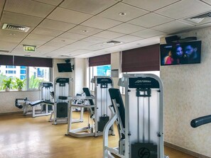 Uma academia com diversos equipamentos de exercício, uma tela de televisão e grandes janelas.