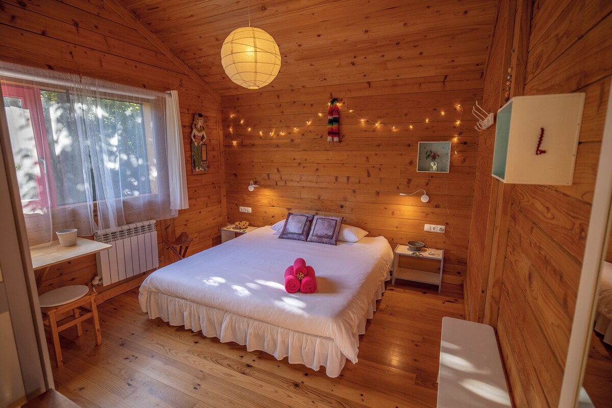 Quarto de cabana de madeira com cama, mesas de cabeceira, janela com cortinas, luminária pendente e um pequeno objeto decorativo na parede.