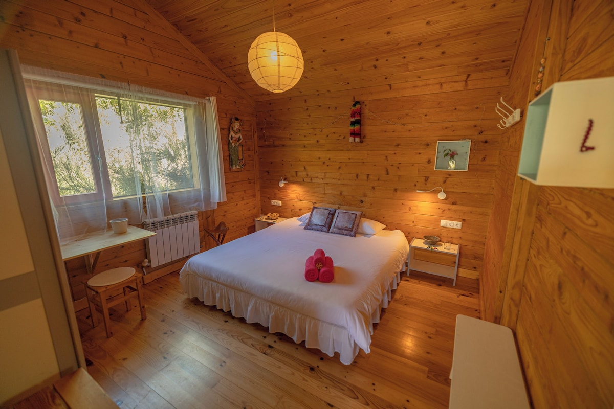 Quarto de cabana de madeira com cama, mesas de cabeceira, janela e luminária pendente.