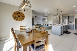 Cozinha moderna com armários cinza, sala de jantar com mesa e cadeiras de madeira, e sala de estar com sofá e um relógio de parede grande.