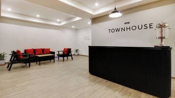 Uma área de recepção com balcão preto, espaço de espera com cadeiras pretas e almofadas vermelhas, e um letreiro fixado na parede com a inscrição 'OYO TOWNHOUSE'.