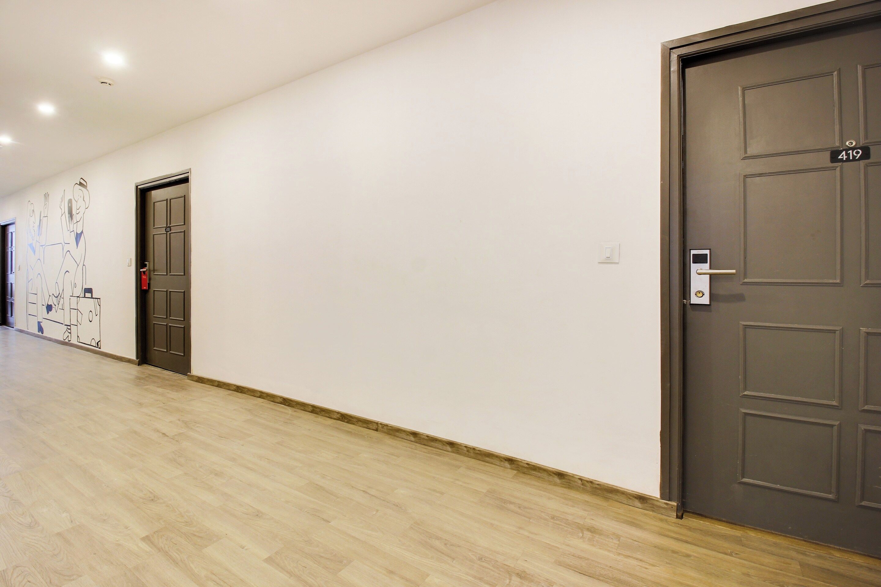 Corredor com piso de madeira, porta escura com placa de número e parede com um desenho linear simples.
