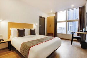 Quarto de hotel com uma cama grande, escrivaninha, cadeira, guarda-roupa e janela com cortinas.