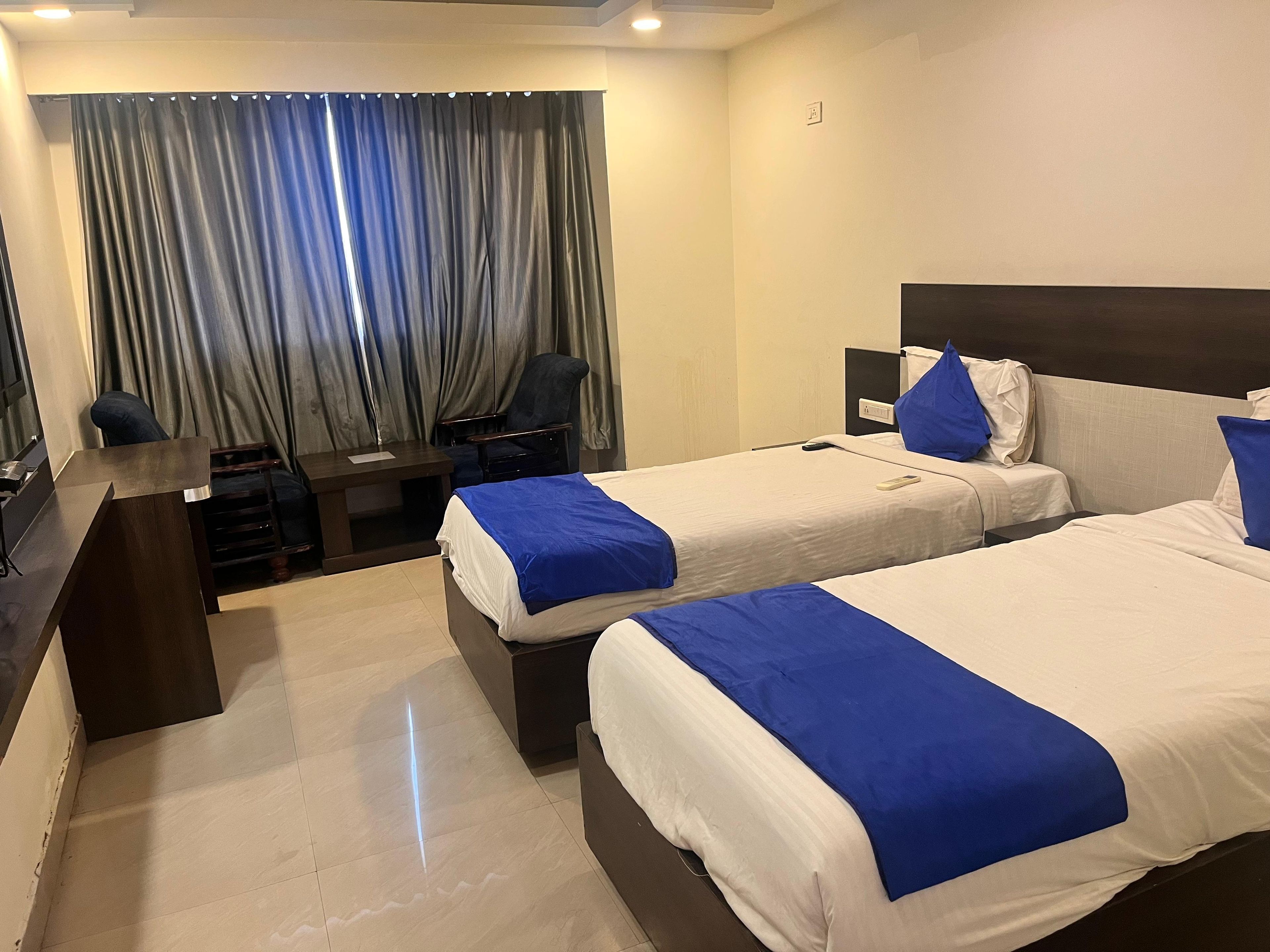 Quarto de hotel com duas camas, uma escrivaninha, uma cadeira e uma janela grande com cortinas.