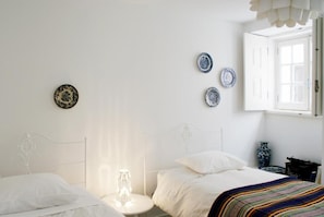 Quarto com estrutura de cama de metal branca, duas almofadas brancas, manta listrada colorida, mesa de cabeceira com abajur e três pratos decorativos na parede.