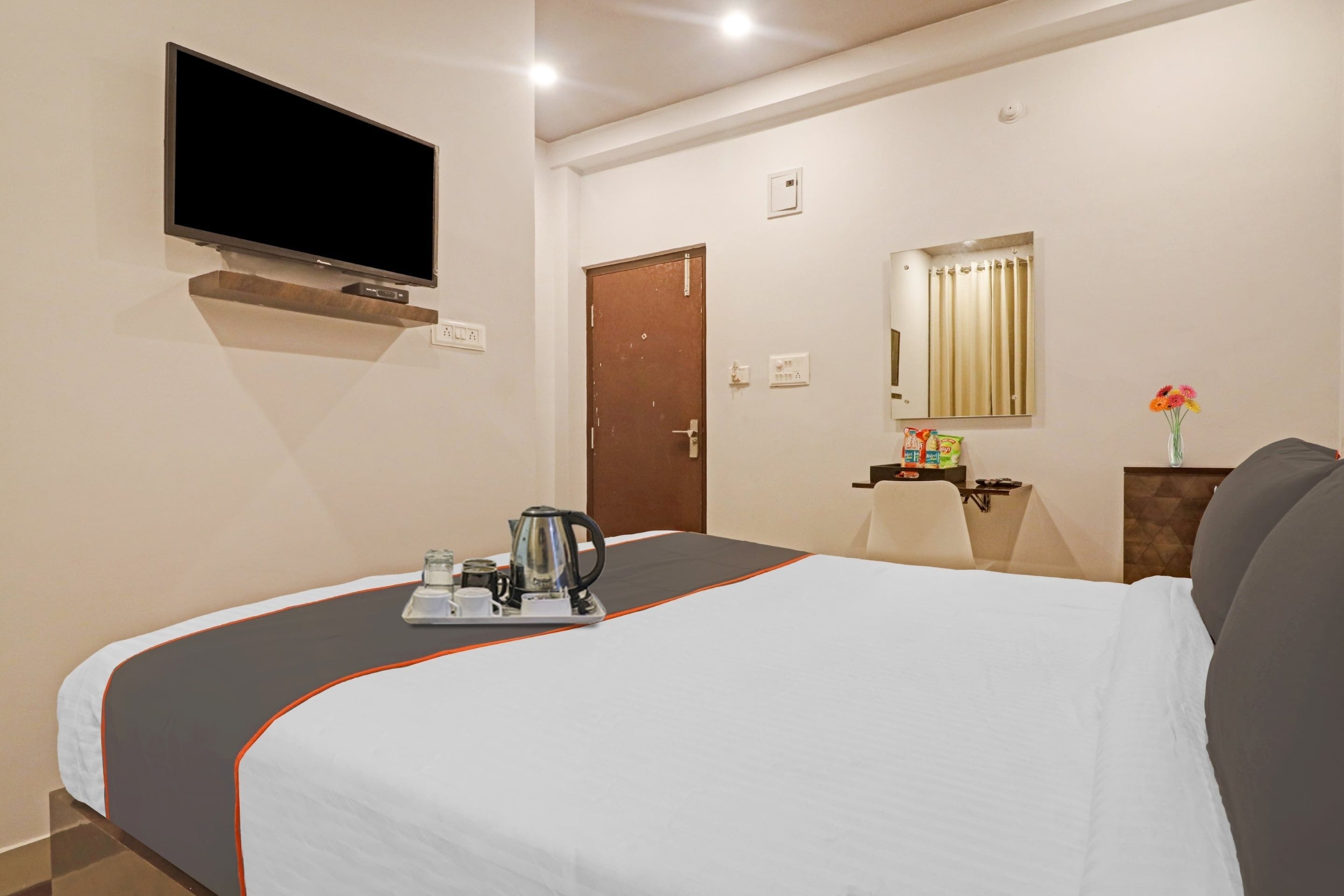 Quarto de hotel com cama, televisão na parede, uma mesinha com chaleira, cadeira e espelho.