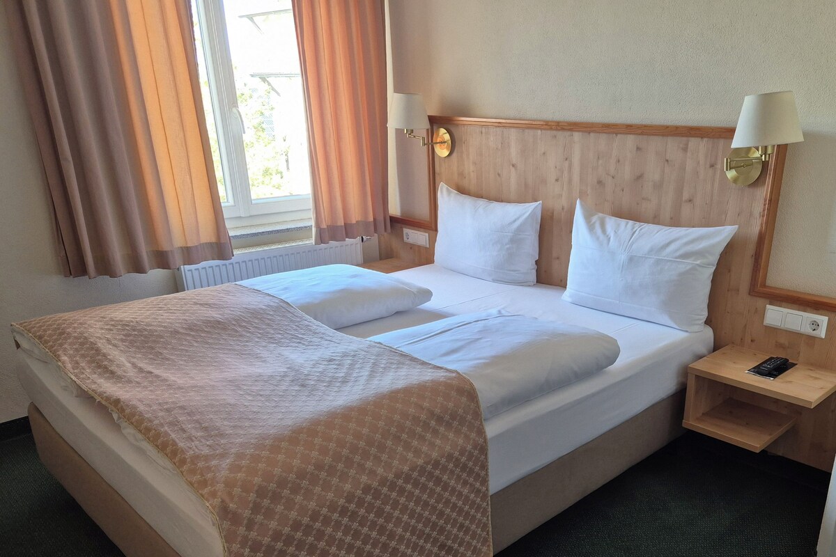 Uma cama de casal com colcha estampada, duas mesas de cabeceira com abajures, e vista para o exterior pela janela.