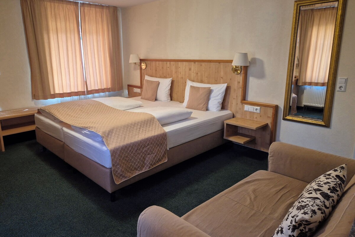 Quarto de hotel com cama, mesas de cabeceira, uma cadeira, espelho e cortinas.