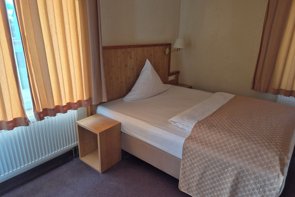 Quarto de hotel com cama, mesa de cabeceira, luminária e janela com cortinas.