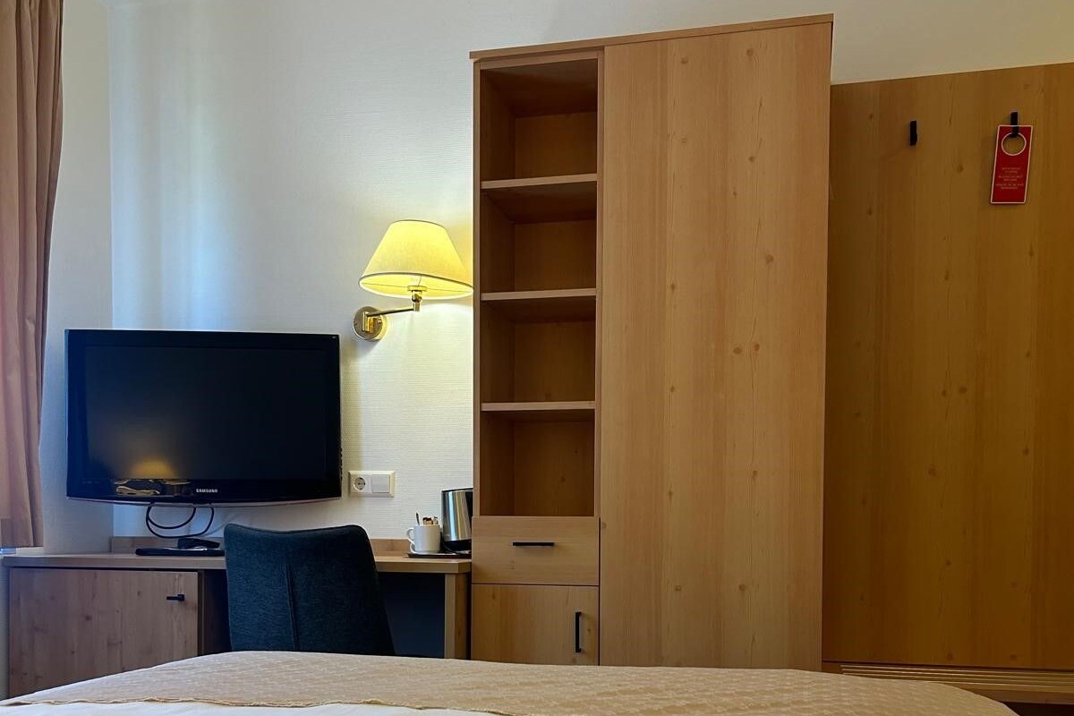 Quarto de hotel com cama, escrivaninha, luminária, televisão e guarda-roupa.