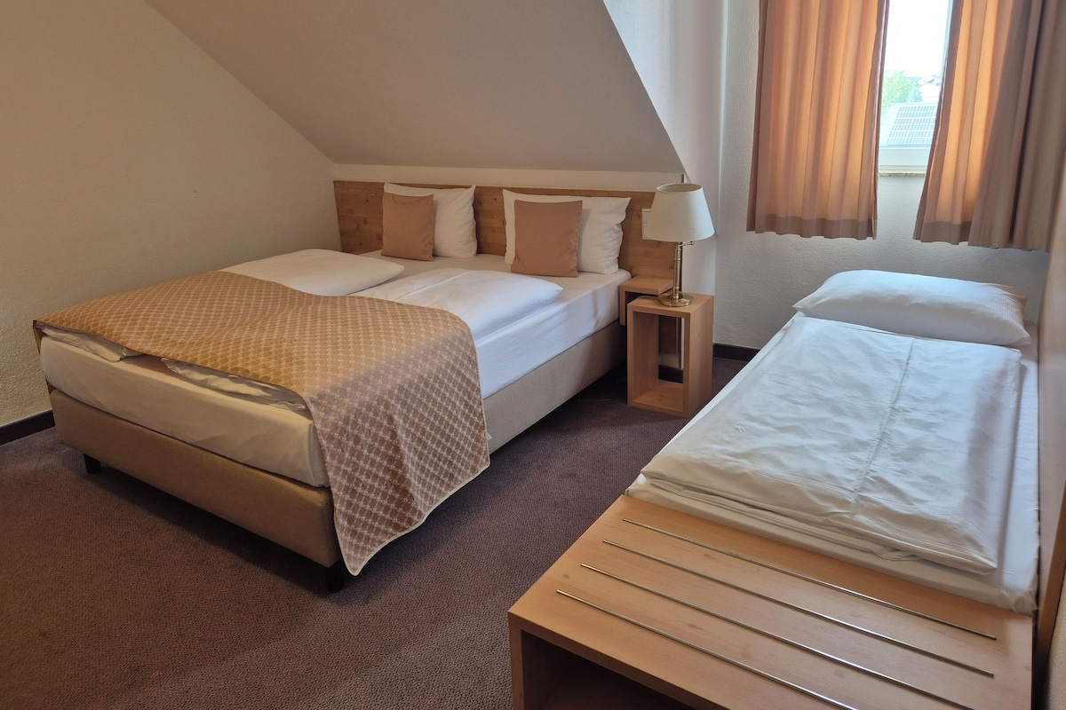 Quarto de hotel com cama, mesa de cabeceira, luminária e janela com cortinas.