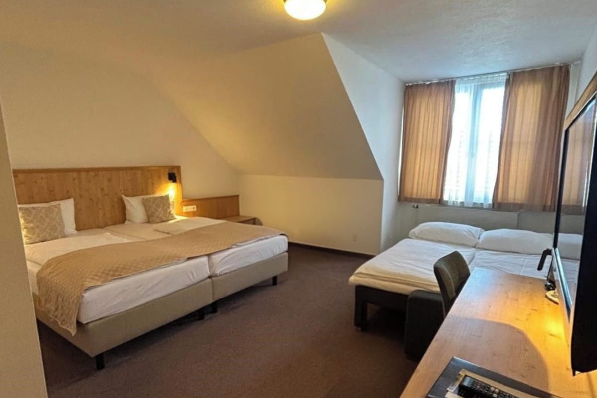 Quarto de hotel com uma cama, uma escrivaninha e uma cadeira. Há uma luminária no teto e uma janela com cortinas.