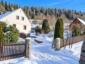 Área residencial coberta de neve, com casas, cerca de madeira e portão de pedra.