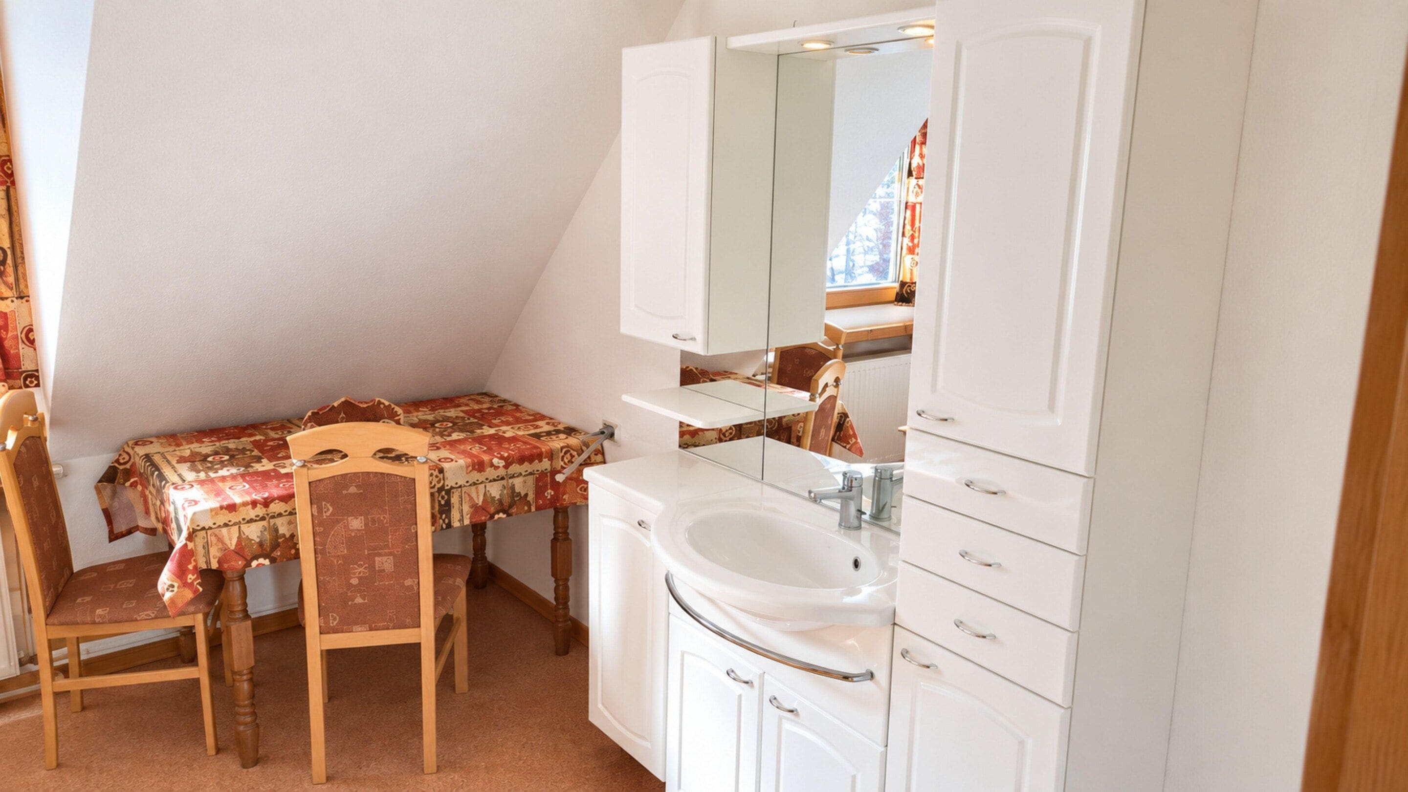 Um quarto com cama, uma cadeira, uma mesa, um espelho e um armário branco.