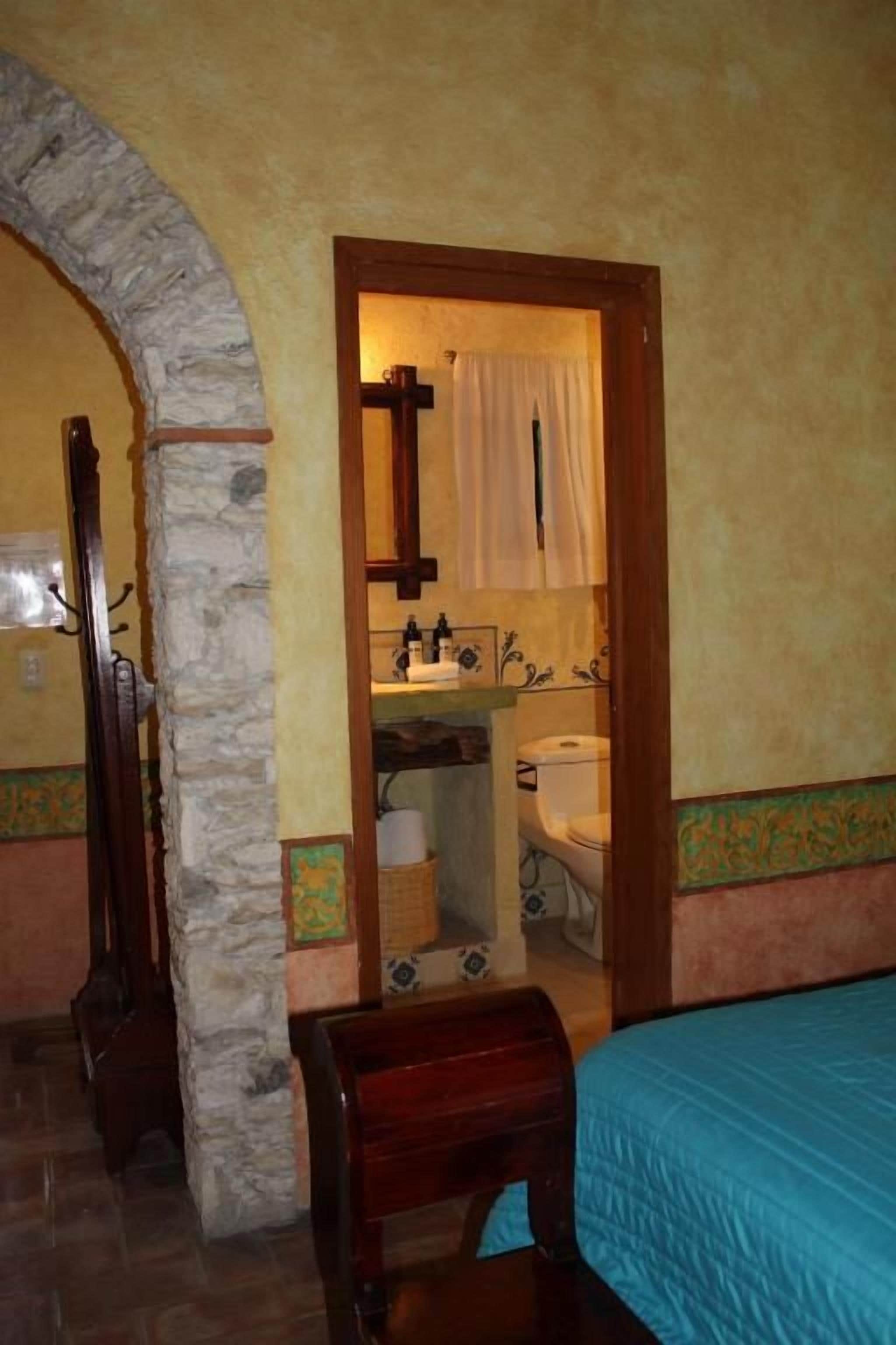 Quarto com arco de pedra que dá para um banheiro, uma cadeira de madeira e uma cama com cobertura azul.