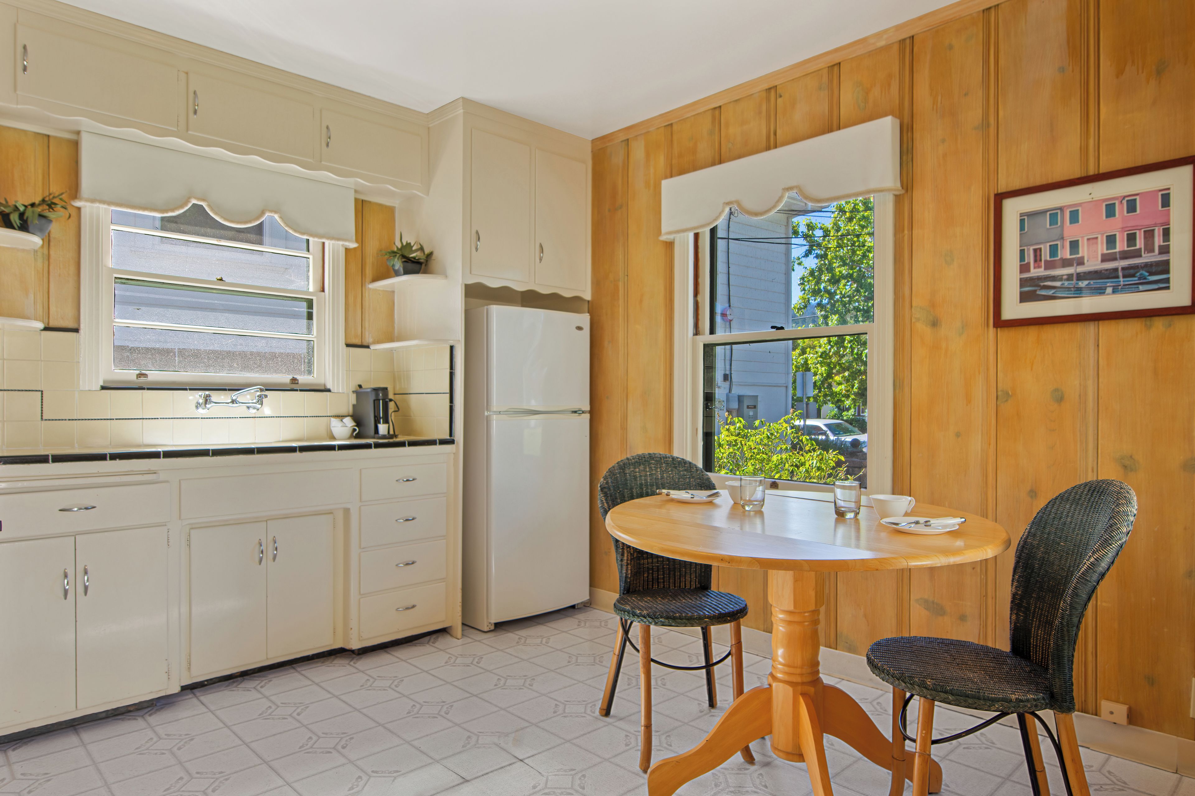 Cozinha com mesa e cadeiras, geladeira, armários e janela com vista para vegetação e um prédio.